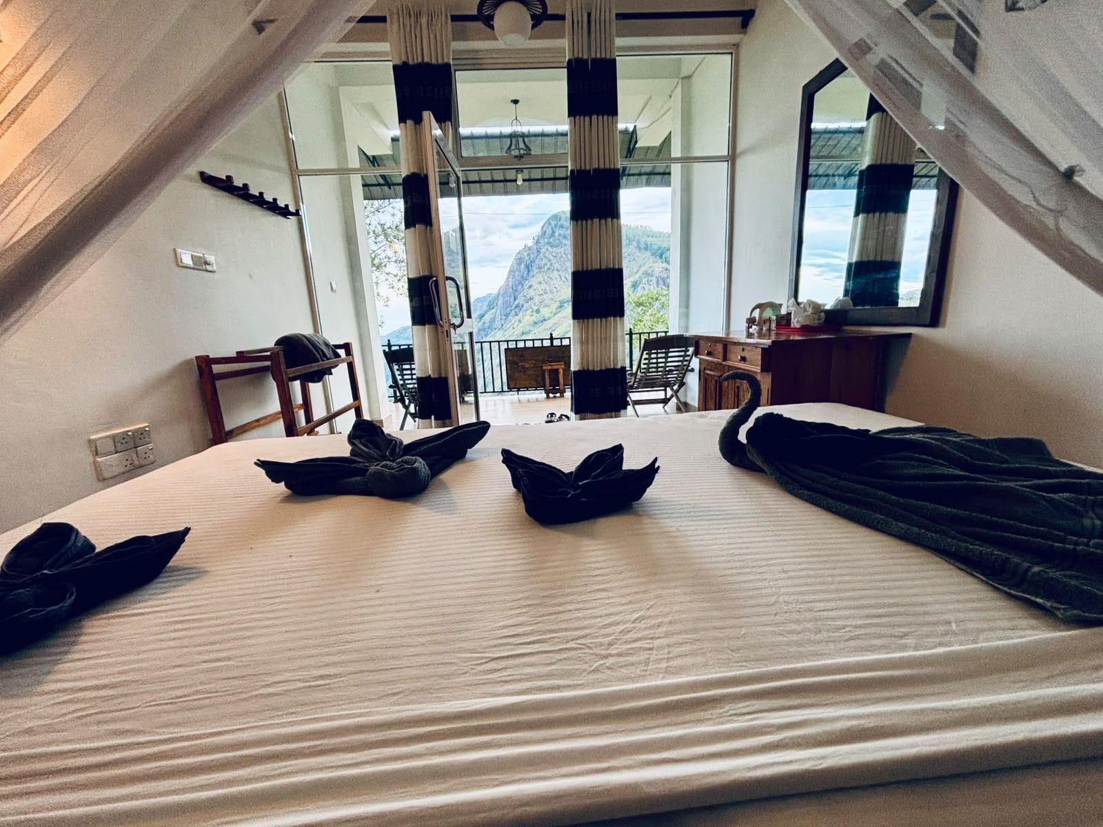 Quarto com cama, escrivaninha, cadeira e vista para montanhas através de amplas janelas.