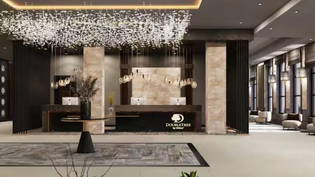 Um hall de hotel moderno com um balcão de recepção central, um lustre decorativo e uma área de estar com sofás.