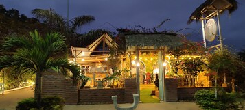 Um edifício rústico com telhado de palha, rodeado por vegetação exuberante e palmeiras.