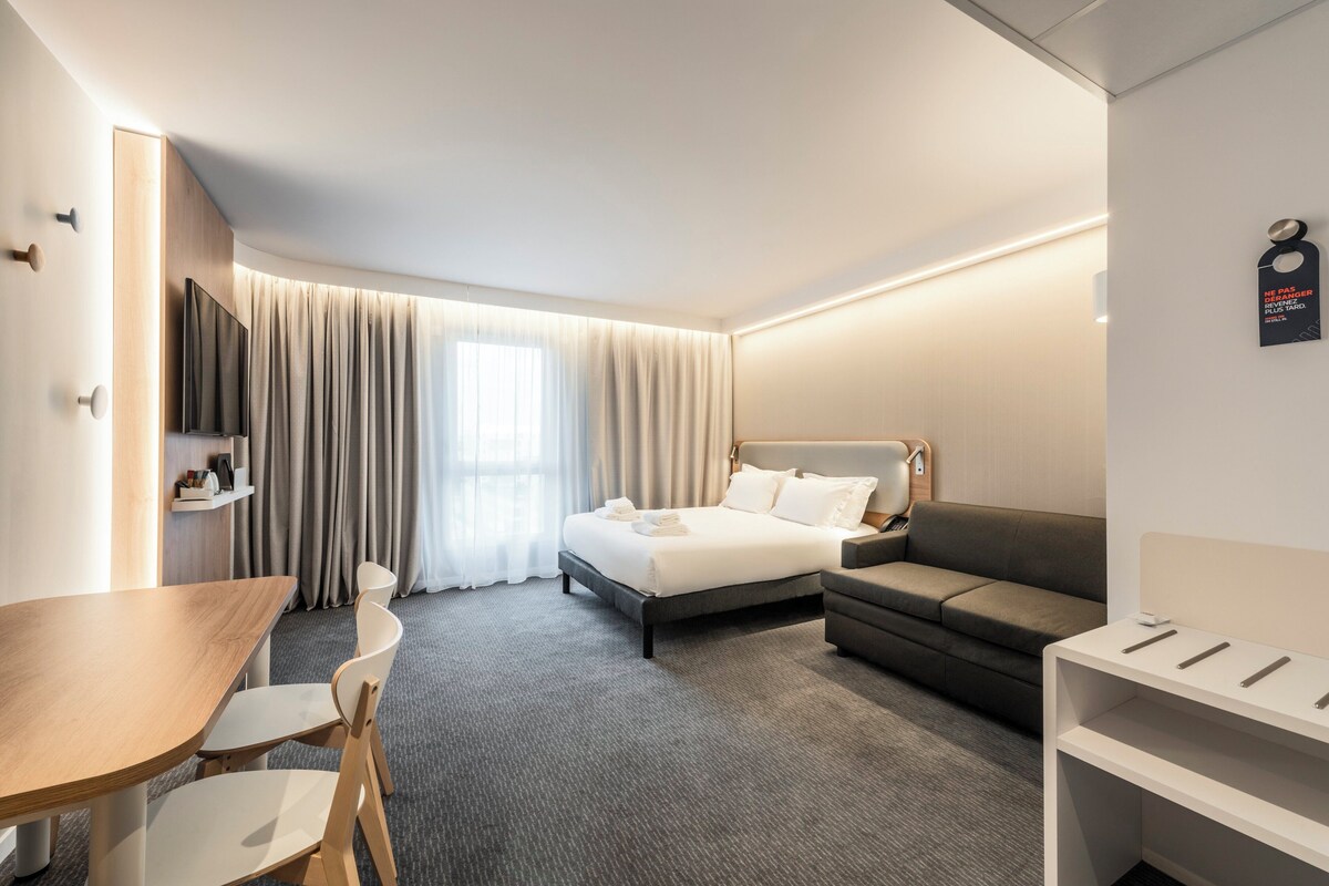 Quarto de hotel com cama, sofá, televisão, escrivaninha e cadeira.