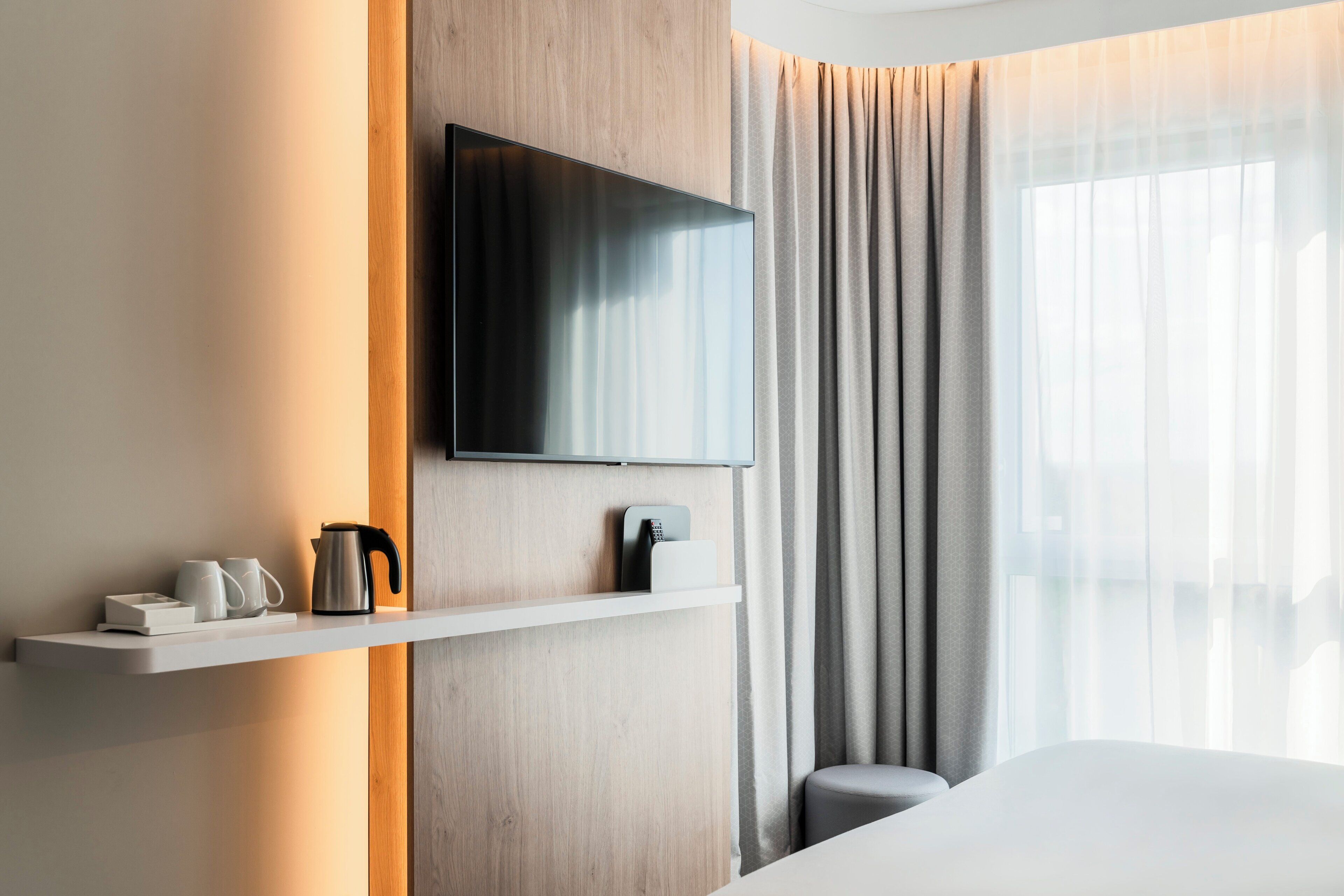 Quarto de hotel moderno com televisão de tela plana fixada em um painel de madeira, uma prateleira branca com cafeteira e xícaras, e uma grande janela com cortinas leves.