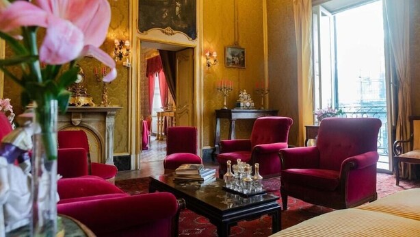Quarto luxuoso com poltronas de veludo vermelho, uma mesa de centro de mármore e uma grande janela com cortinas.