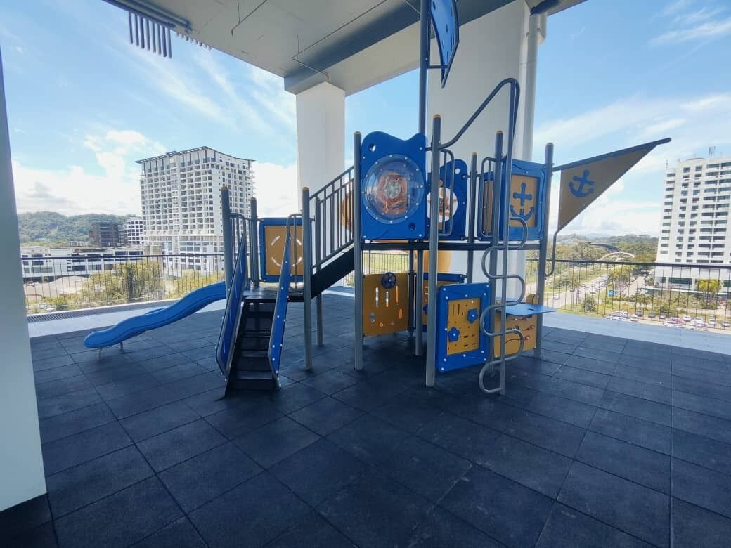 Um parquinho com um escorrega azul e estruturas de escalada, ao fundo edifícios e céu claro.
