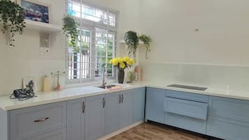 Uma cozinha com armários azul-claros, bancada branca, janela com vista para o verde e um vaso com flores amarelas.