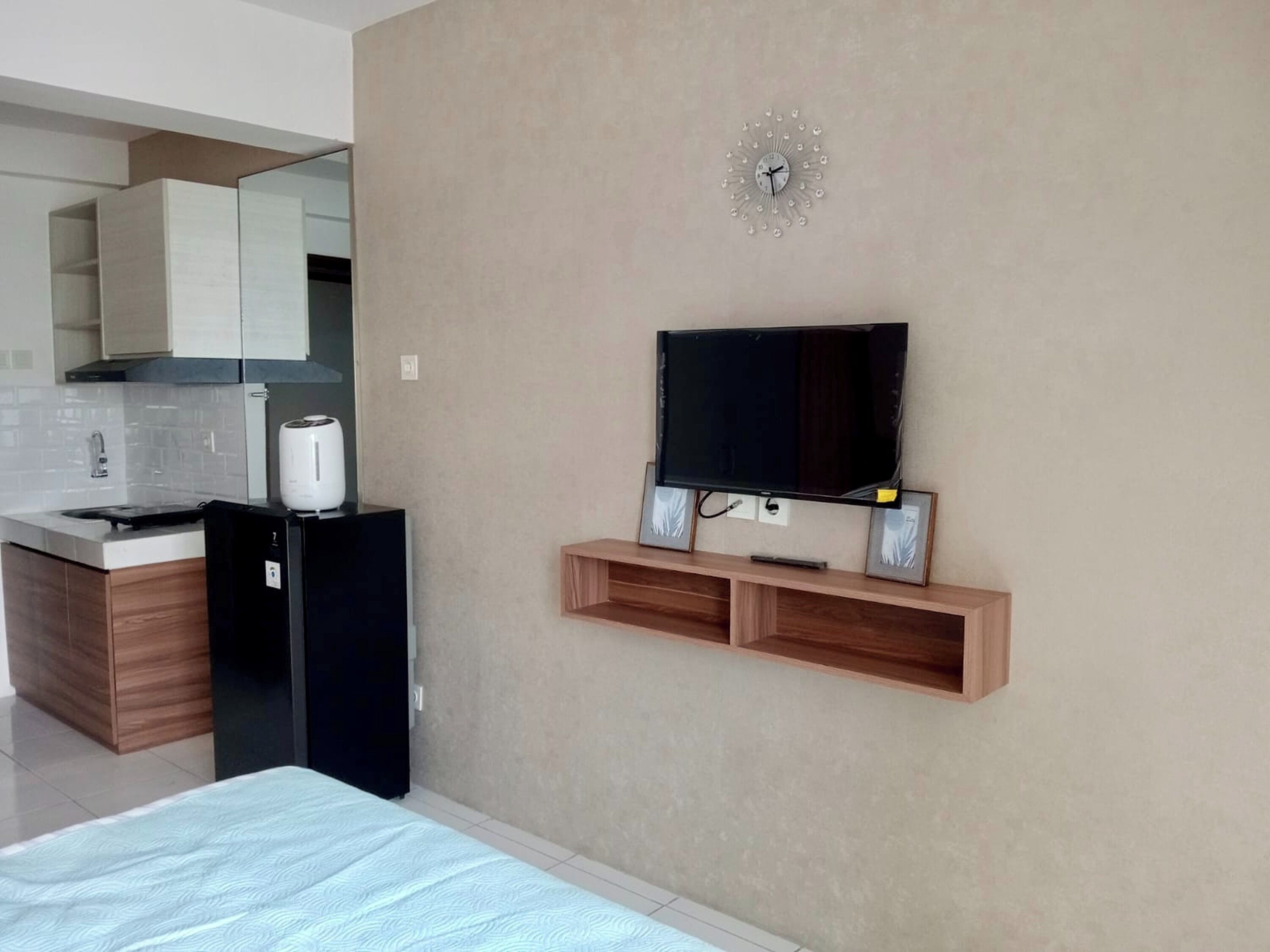 Quarto moderno com cama, televisão fixada na parede, pequena cozinha com frigorífico e máquina de café, e um relógio na parede.
