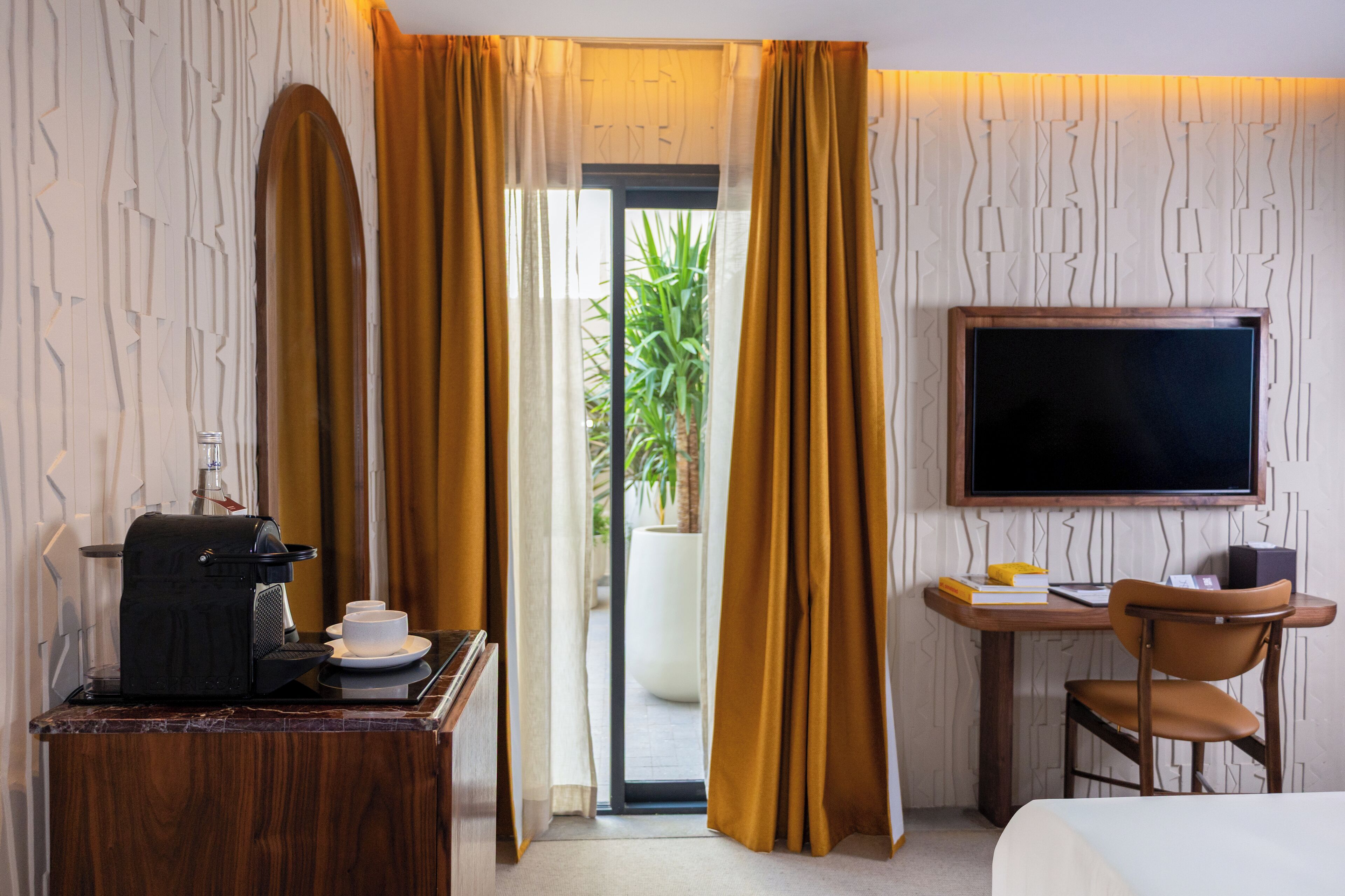 Quarto de hotel com máquina de café, escrivaninha com cadeira, televisão de tela plana e uma grande janela com cortinas.