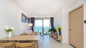 Sala moderna com mesa de jantar de madeira, sofá e televisão. Possui varanda com vista para o mar.