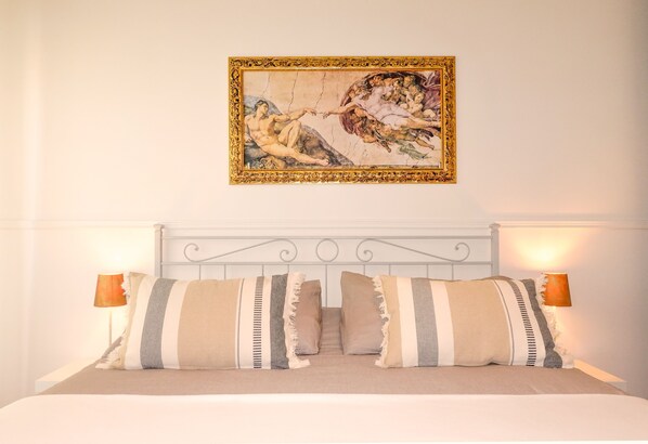 Cama com cabeceira branca, duas luminárias de apoio e um quadro emoldurado acima.