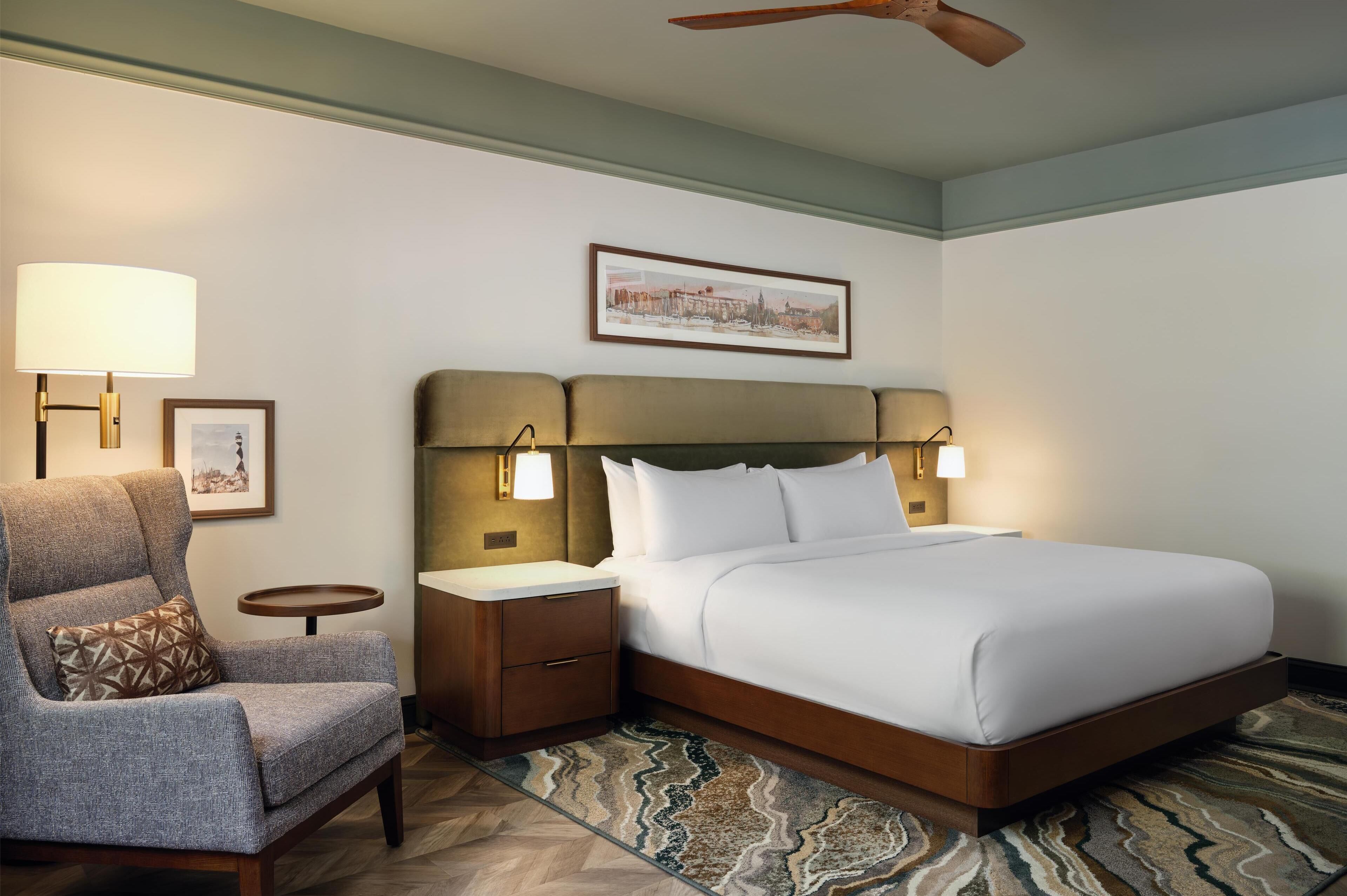 Quarto de hotel com cama, uma cadeira, mesa de cabeceira, abajur e ventilador de teto.