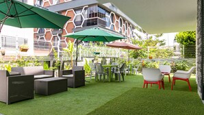 Pátio externo com móveis de vime, cadeiras verdes e vermelhas, e guarda-sóis.