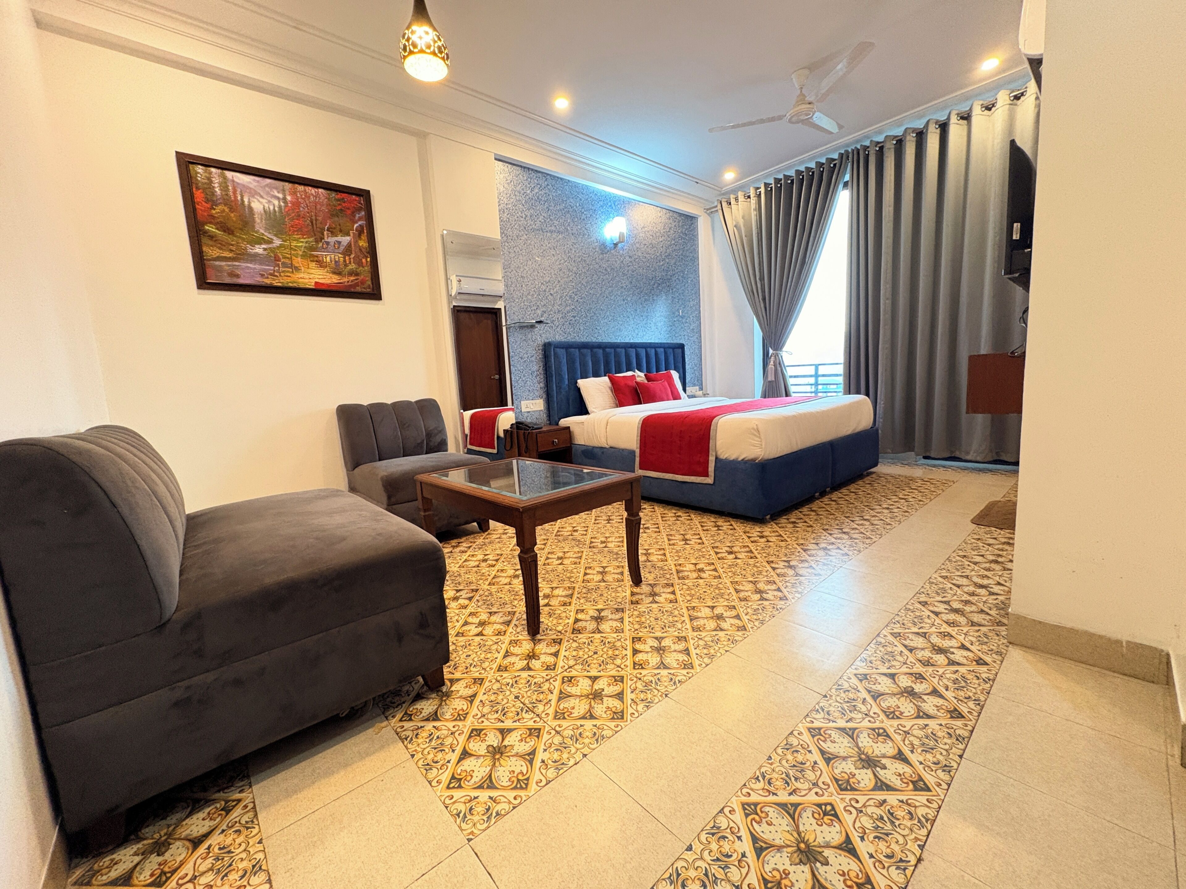 Quarto de hotel com sofá, cama com manta vermelha, tapete estampado, quadro na parede e ventoinha no teto.