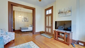 Uma sala de estar com piso de madeira, um sofá, uma televisão e uma porta que dá para outro quarto.