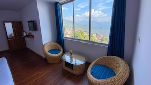 Quarto de hotel com uma janela grande, duas cadeiras de vime com almofadas azuis, uma mesa pequena com um objeto decorativo, uma cama e uma televisão na parede.