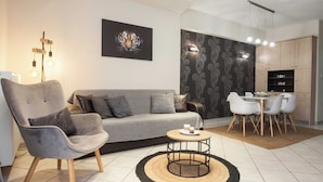 Uma sala de estar moderna com um sofá cinzento, uma poltrona cinzenta, uma mesa de centro redonda pequena e uma área de jantar com mesa e cadeiras brancas.