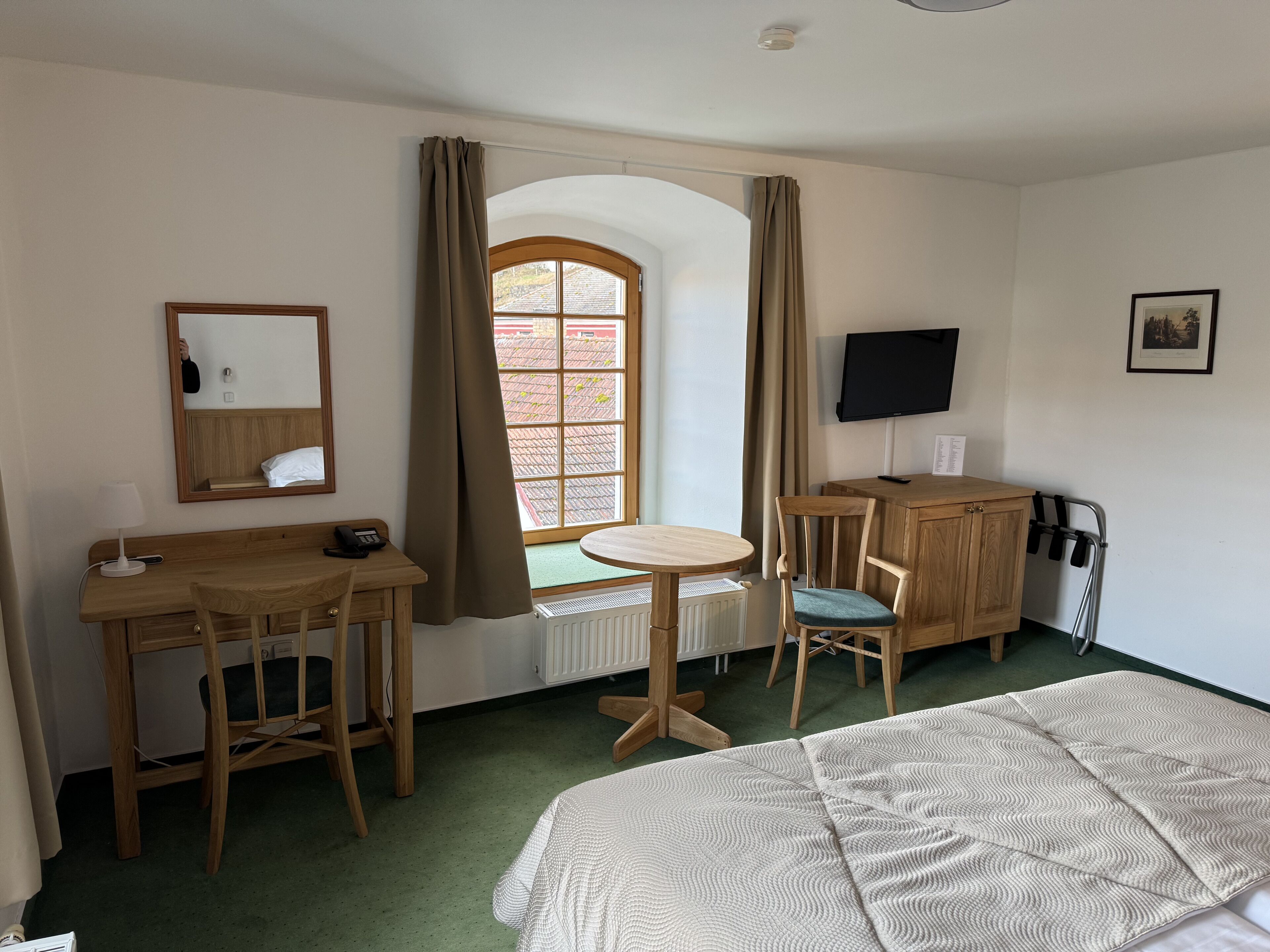 Quarto de hotel com cama, escrivaninha com cadeira, mesinha, televisão e janela com cortinas.