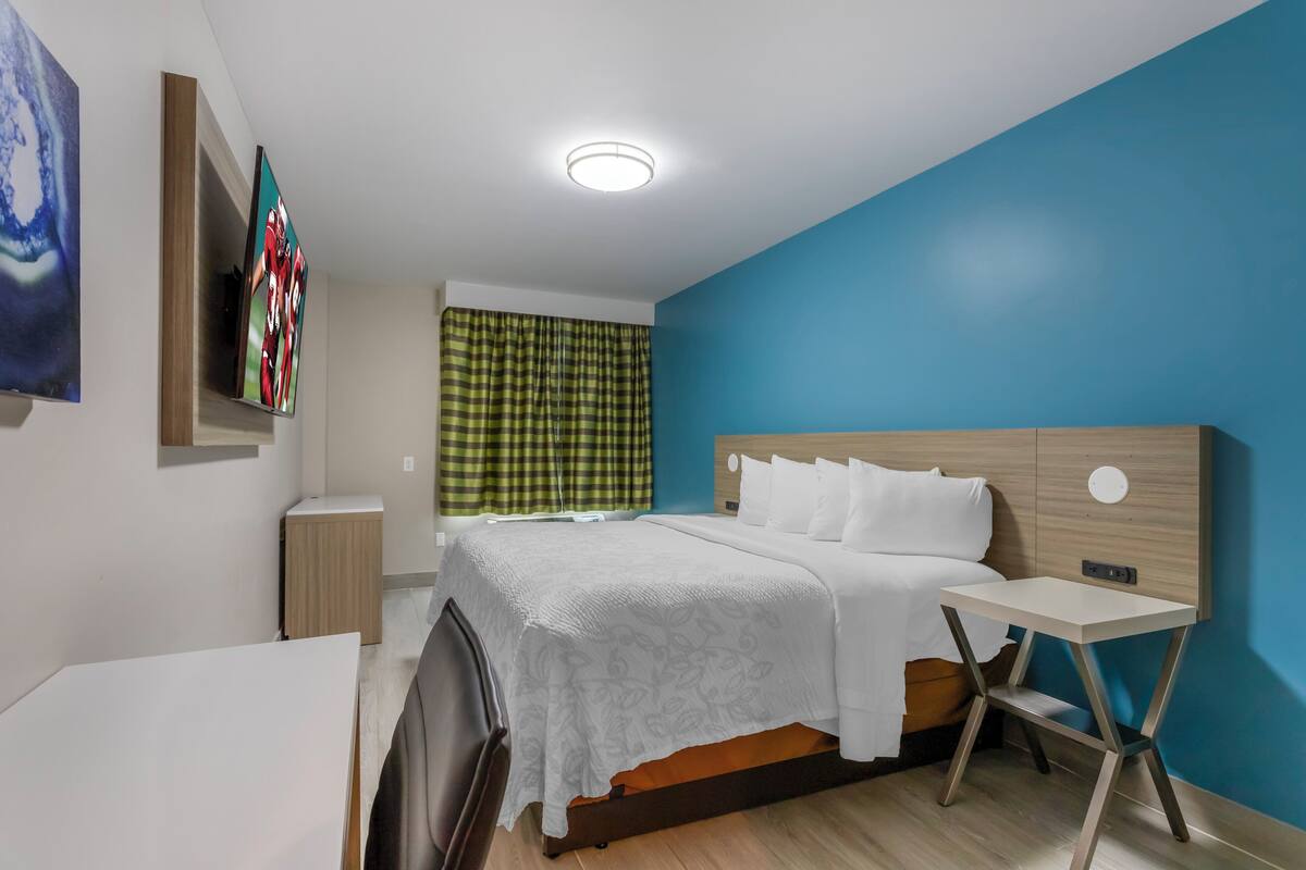 Quarto de hotel moderno com cama, mesa de cabeceira e televisão fixada na parede.