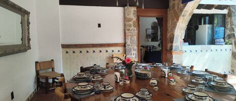 Uma sala de jantar com uma grande mesa de madeira posta para uma refeição, rodeada por cadeiras.