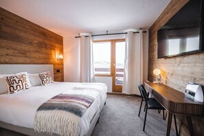 Quarto de hotel com cama, escrivaninha, televisão e janela com cortinas.