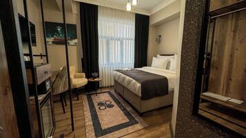 Quarto de hotel com cama, escrivaninha, cadeira, mesinha com um vaso e janela com cortinas.