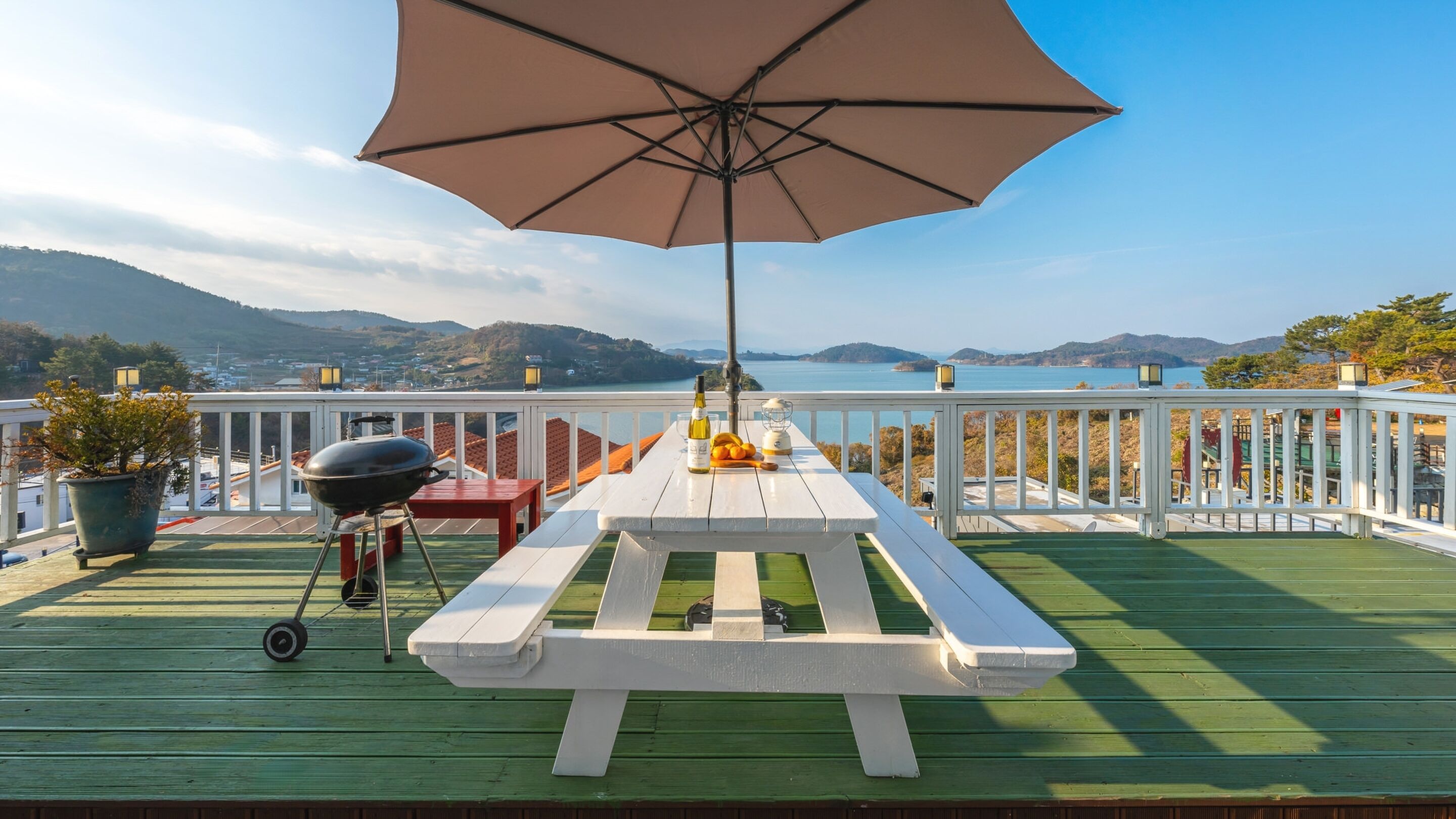 Um terraço com mesa para piquenique, churrasqueira e guarda-sol, com vista para um lago e colinas.
