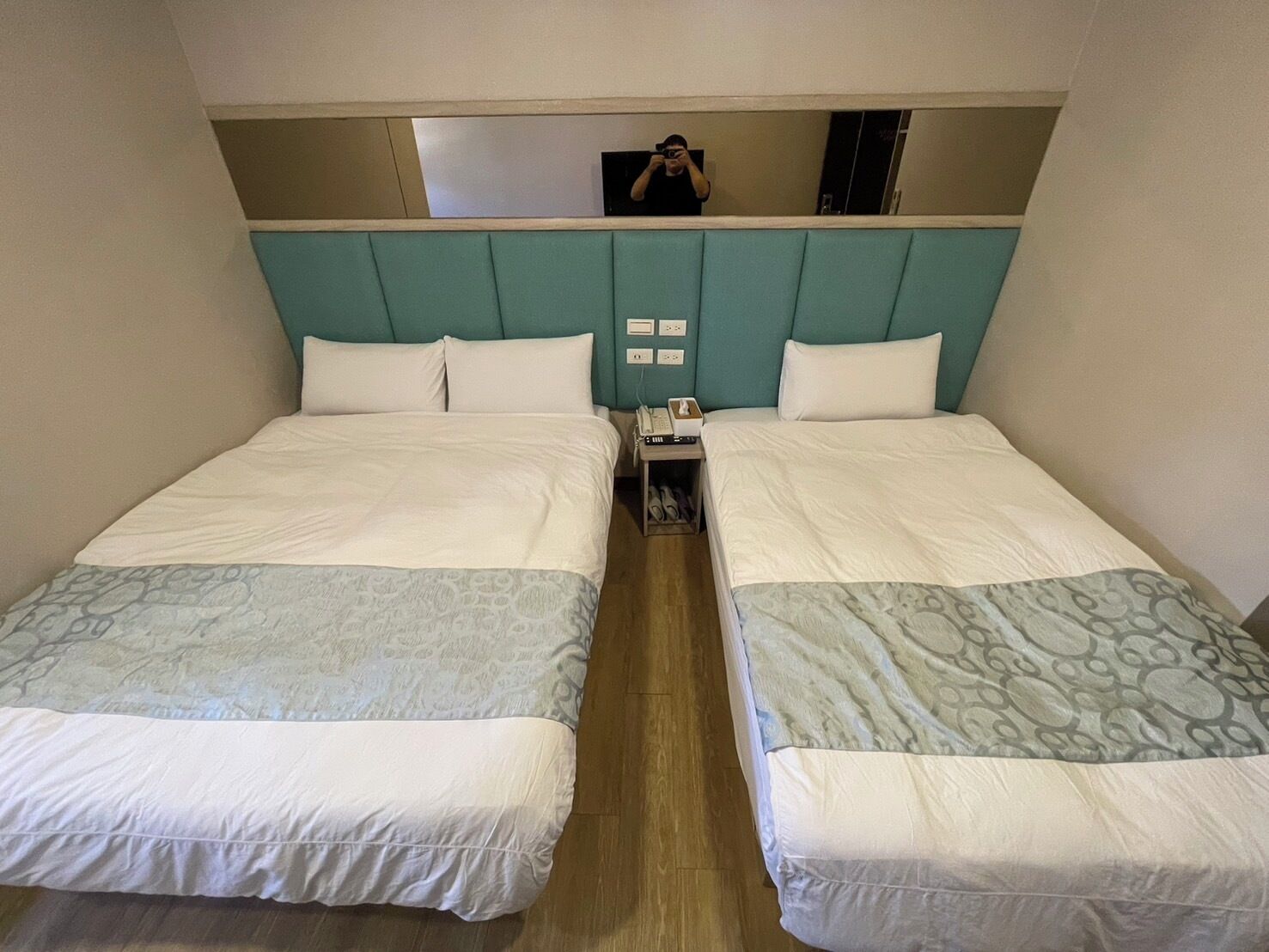 Quarto de hotel com duas camas, uma mesa de cabeceira com abajur e um espelho refletindo uma pessoa.