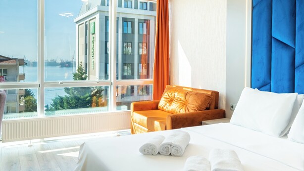 Quarto de hotel com uma janela grande, uma cama com roupa branca, uma poltrona laranja e vista para um edifício à distância, através da água.