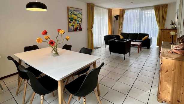Uma sala de jantar com uma mesa branca, cadeiras pretas e um vaso de flores. Há um sofá e um abajur ao fundo.