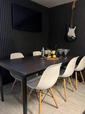 Uma sala de jantar com uma mesa de madeira escura, cadeiras brancas, um violão, um coqueteleira e uma garrafa de cava.