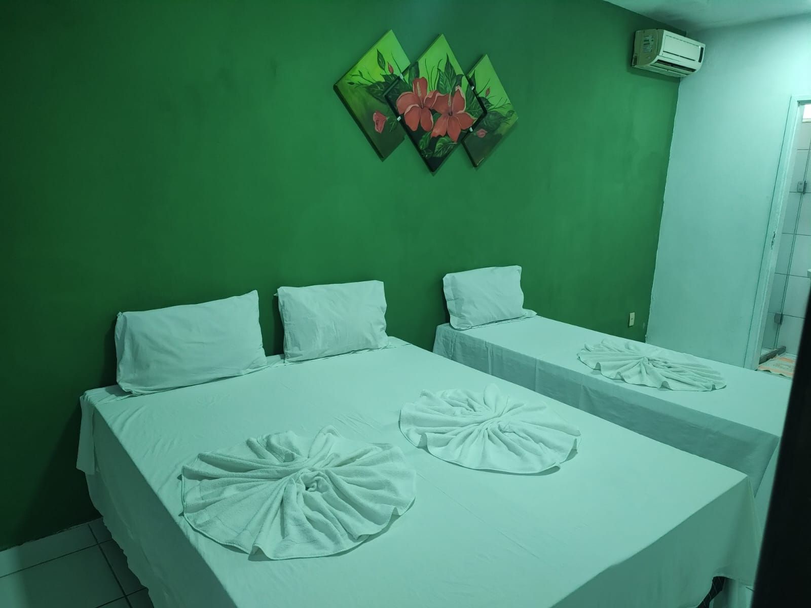 Quarto com duas camas, cada uma com roupa de cama branca e uma toalha sobre a cama. A parede é verde e há uma pintura de flores acima das camas.