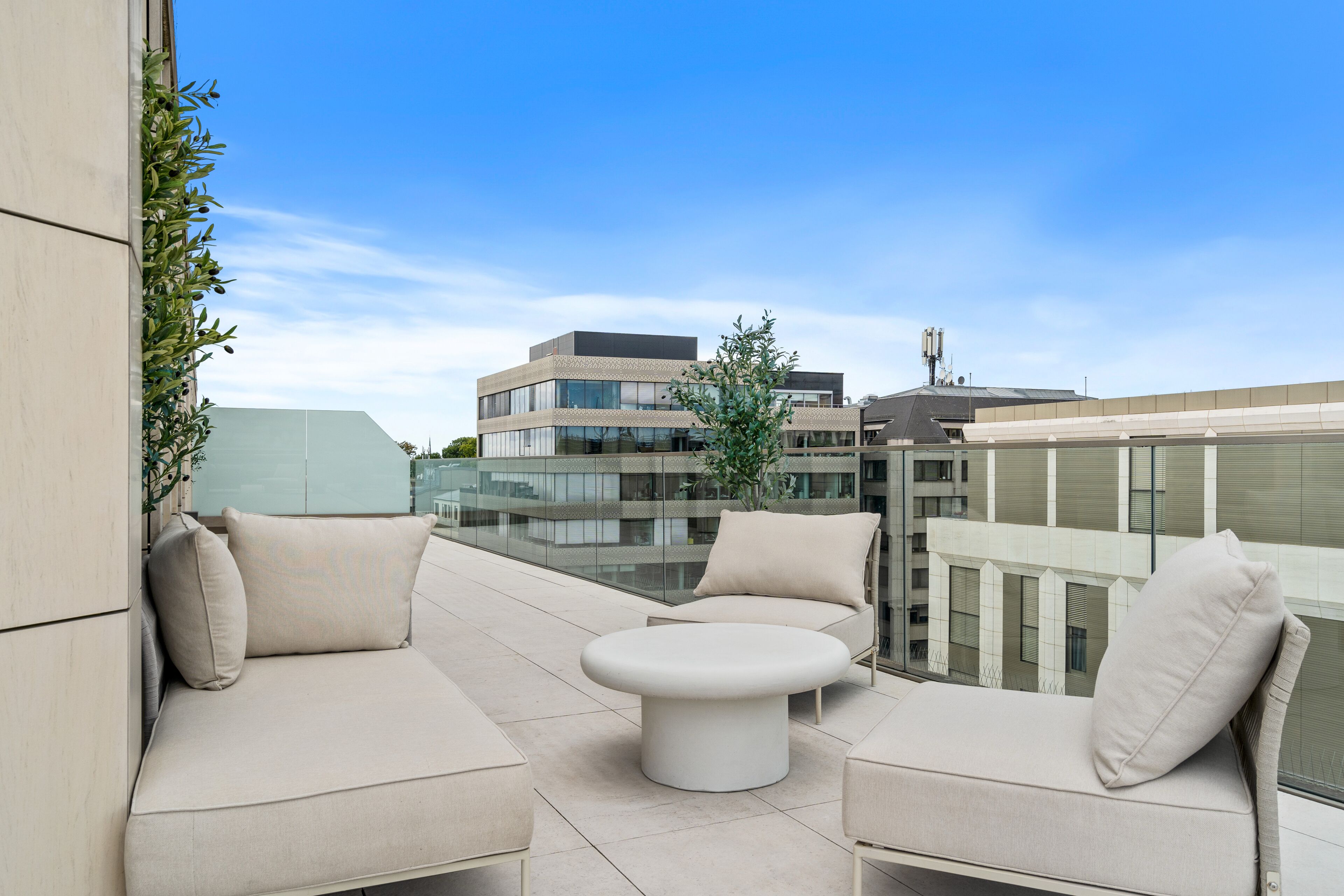 Terraço no telhado com sofá bege, mesa redonda e guarda-corpo de vidro.