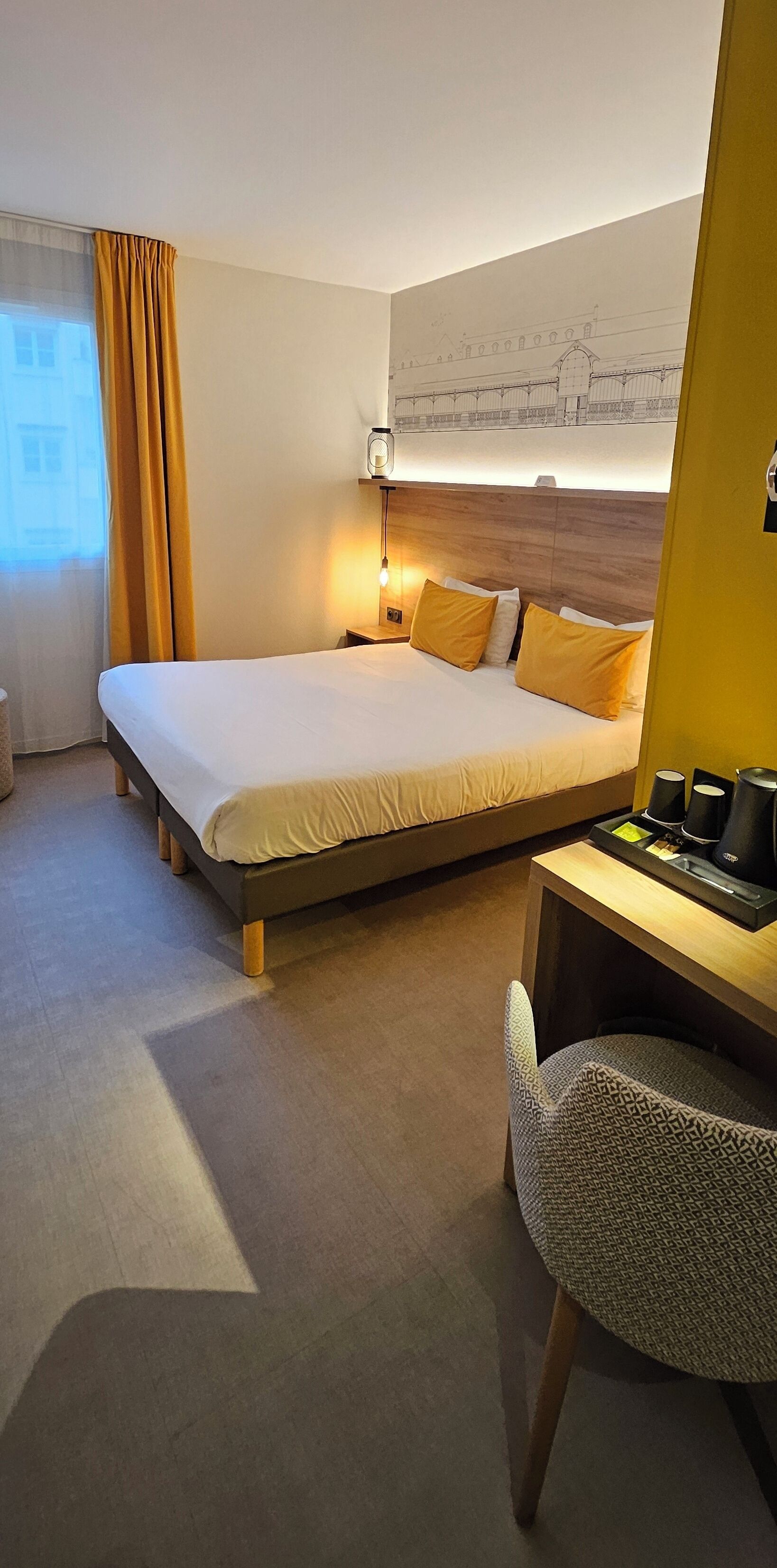 Quarto de hotel com cama, mesa de cabeceira, luminária, cadeira e parede com desenho.