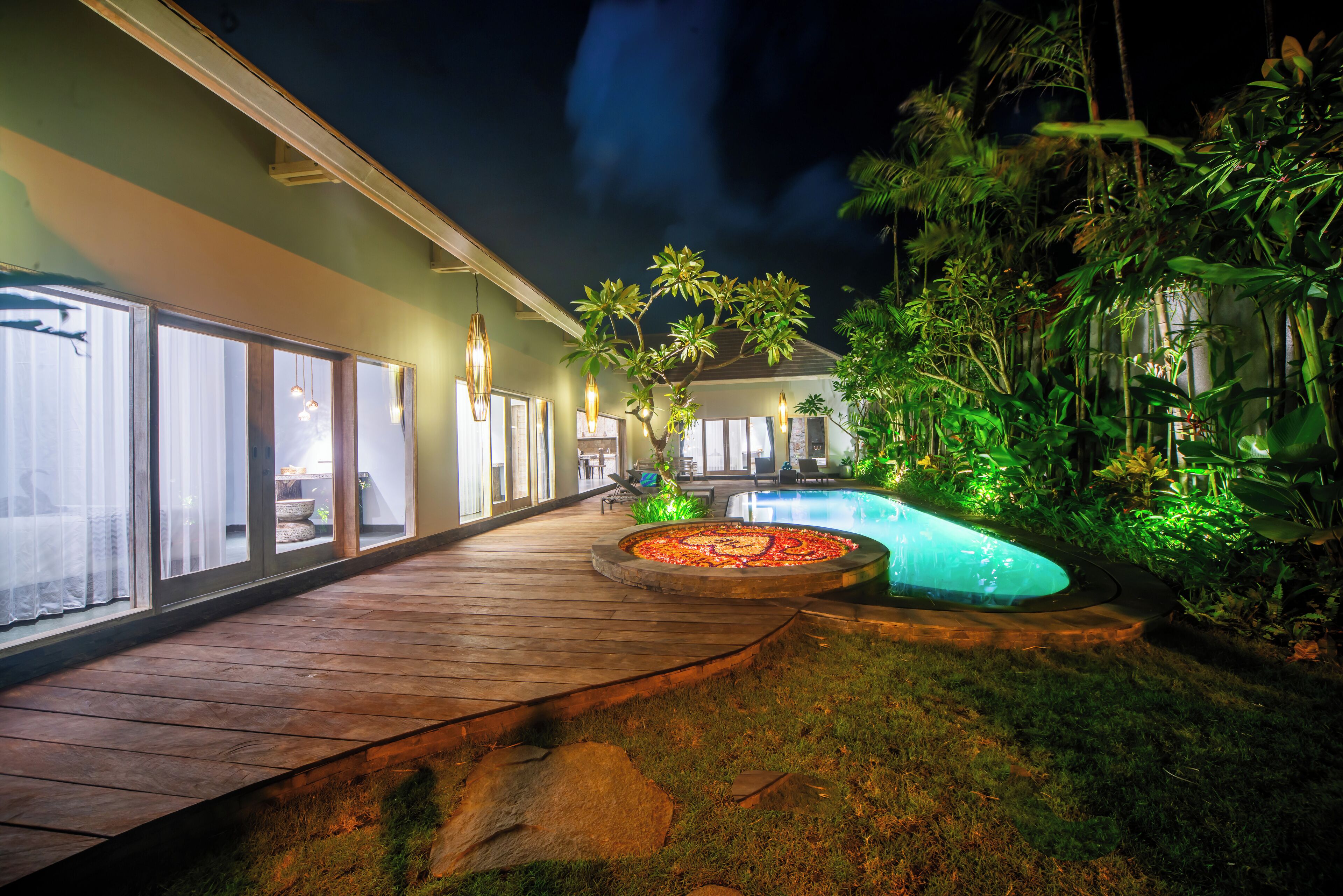 Um deck de madeira leva a uma área de piscina com design de mosaico circular, cercada por vegetação exuberante e um espaço ao ar livre bem iluminado.