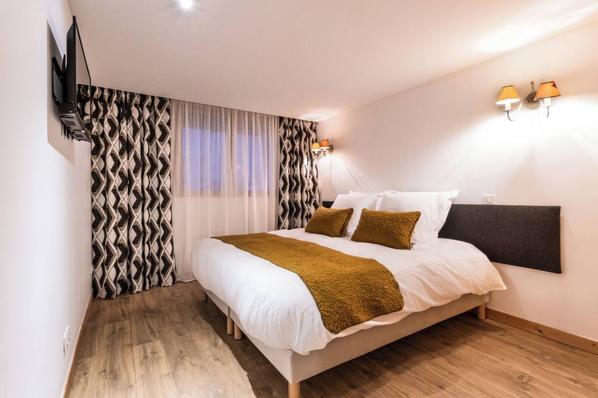 Um quarto com cama, cortinas, TV de tela plana na parede e luminária fixa na parede.