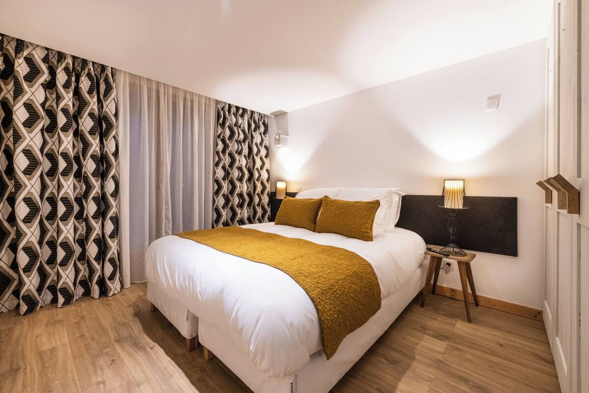 Quarto de hotel com uma cama grande, cabeceira, mesa de cabeceira com abajur e cortinas estampadas.