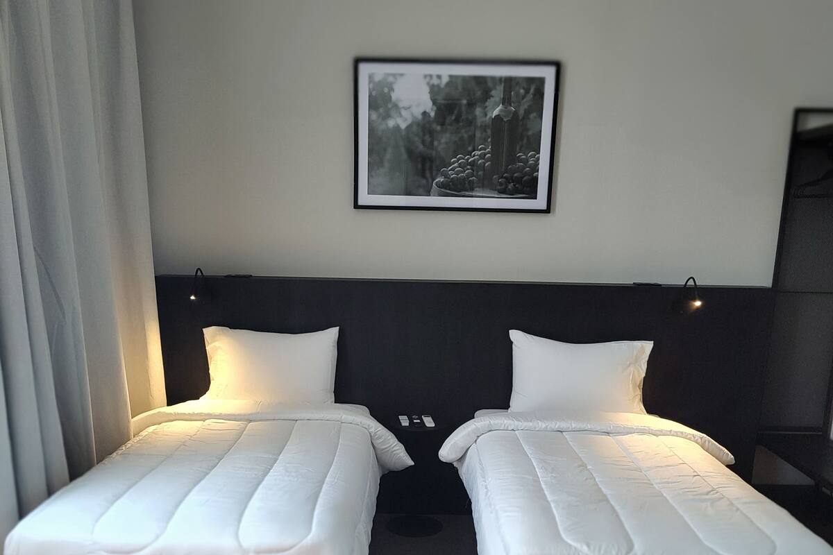 Quarto de hotel com duas camas de solteiro, cabeceira escura, quadro na parede e piso de madeira.