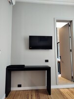 Quarto com televisão de tela plana na parede, mesa de consola preta e porta de acesso a outro espaço.