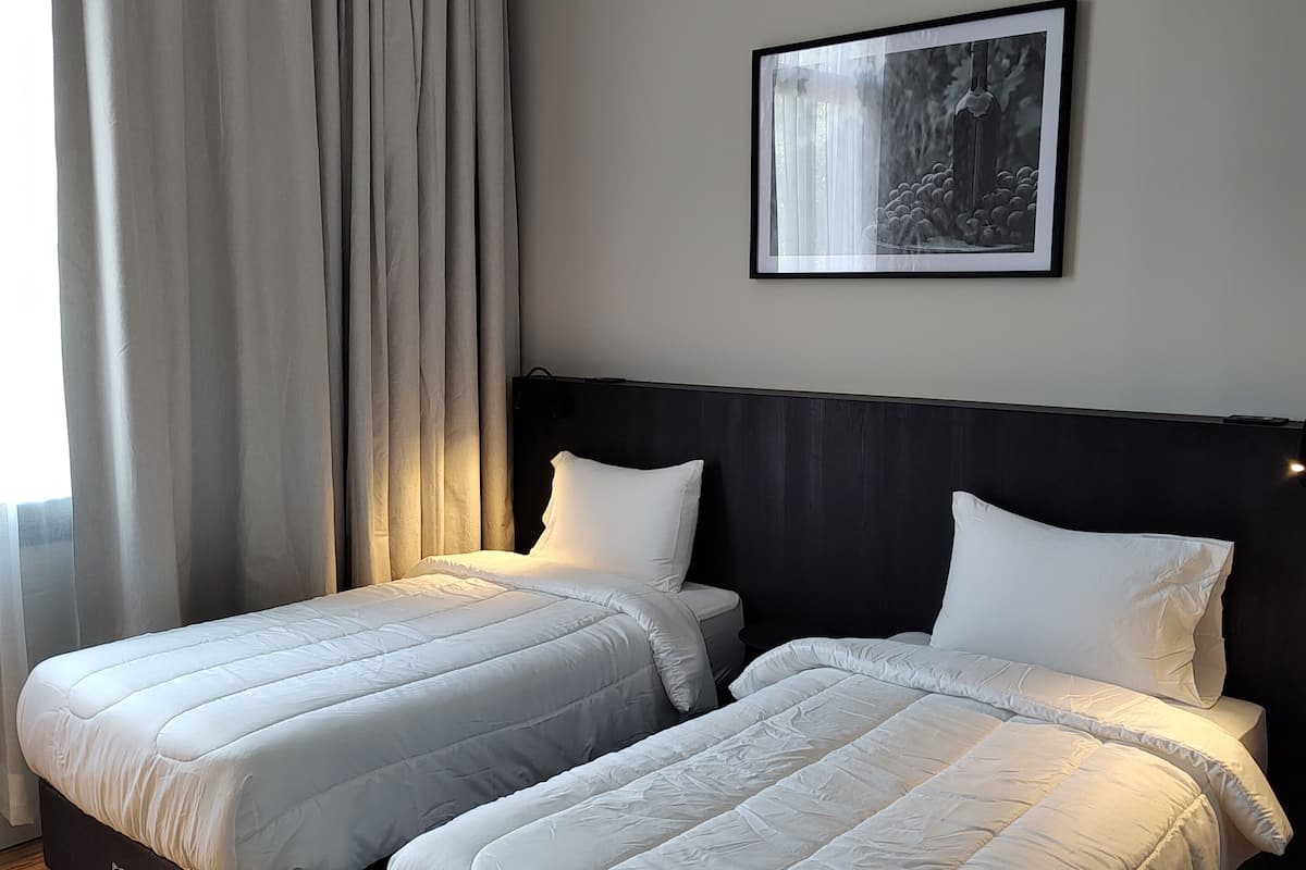 Quarto de hotel com duas camas de solteiro, cabeceira escura, um quadro na parede e uma janela com cortinas.