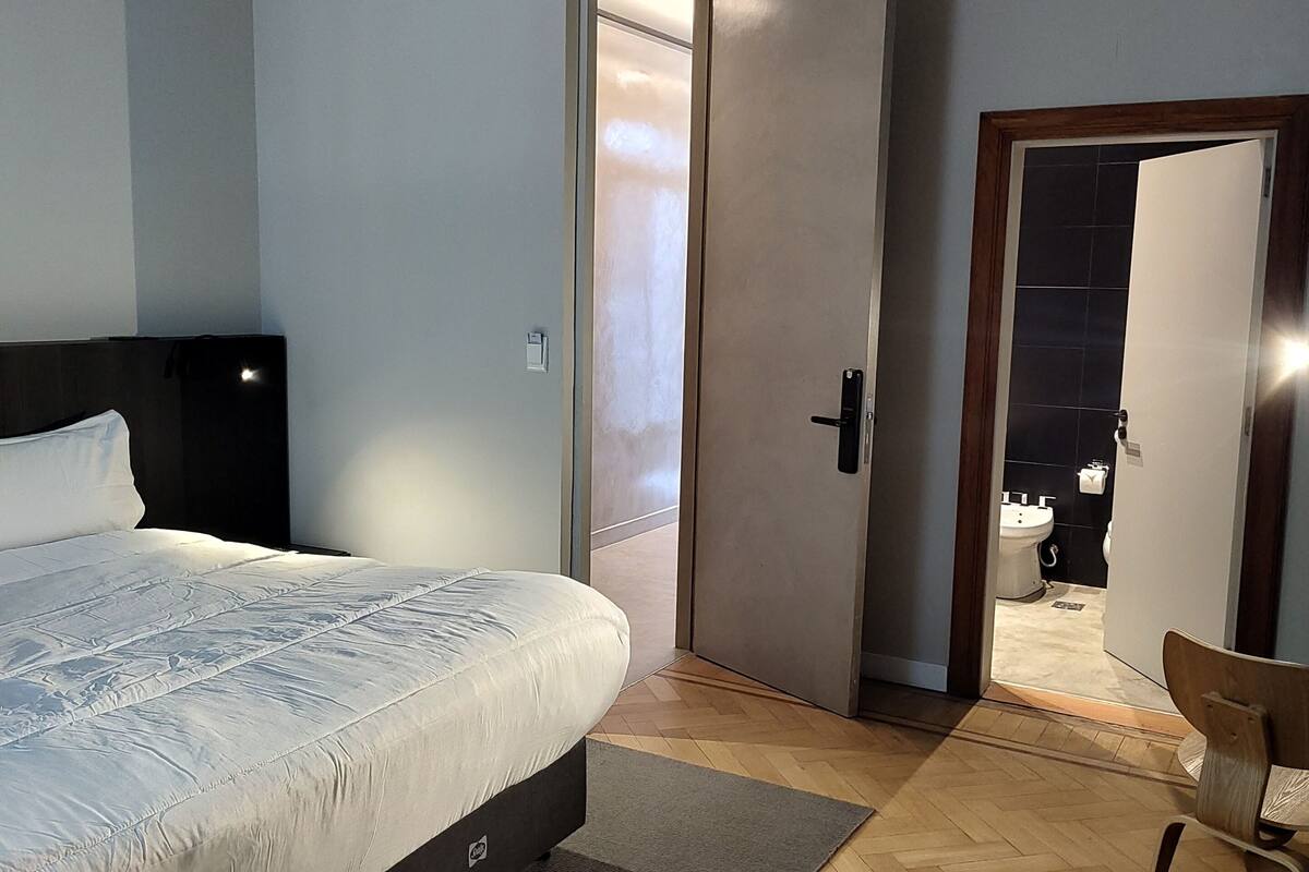 Quarto com cama, uma cadeira, armário e banheiro visível por uma porta aberta.