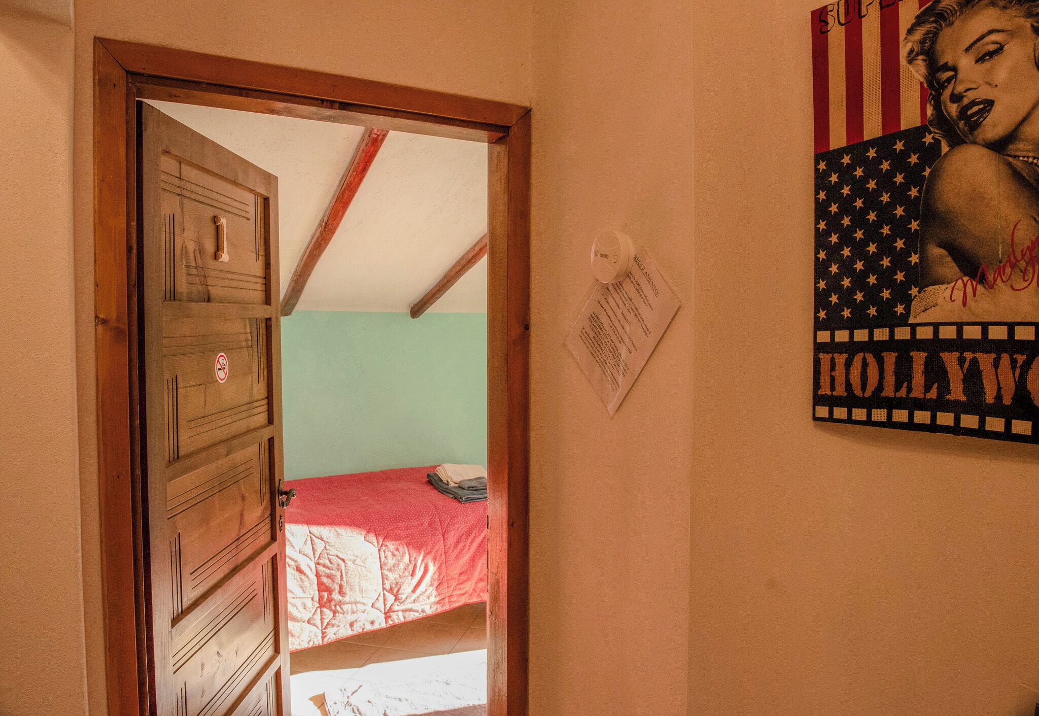 Um quarto com porta de madeira, uma cama com manta vermelha e um cartaz na parede.