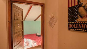 Um quarto com porta de madeira, uma cama com manta vermelha e um cartaz na parede.