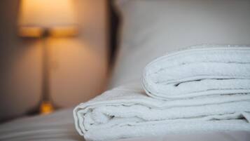 Uma pilha organizada de toalhas brancas sobre uma cama, com uma luminária ao fundo.