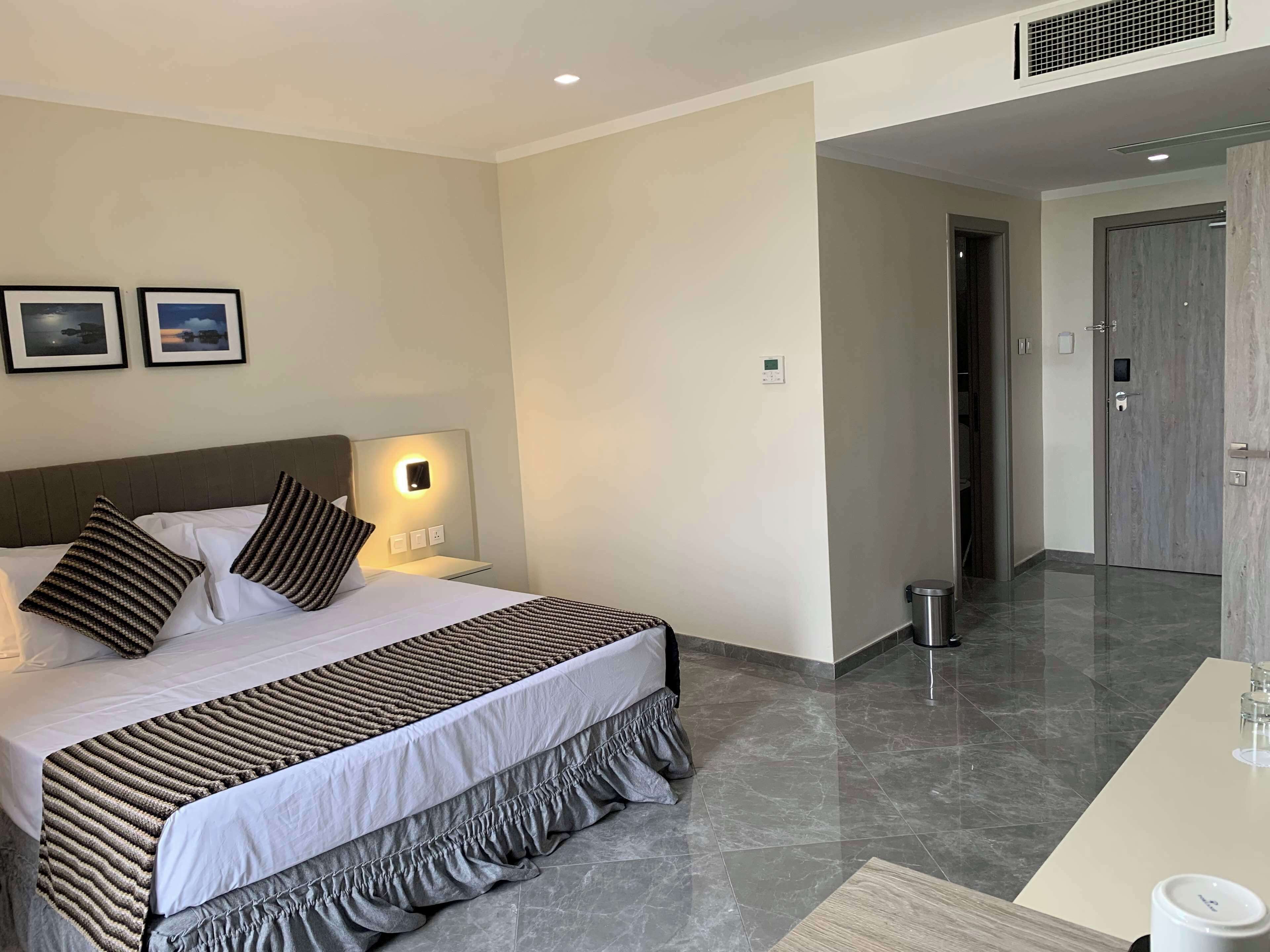 Quarto de hotel com cama, mesa de cabeceira, luminária fixada na parede e porta que dá para outro cômodo.