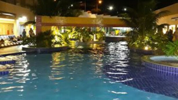 Vista noturna de uma piscina com bordas azulejadas, cercada por edifícios e palmeiras.
