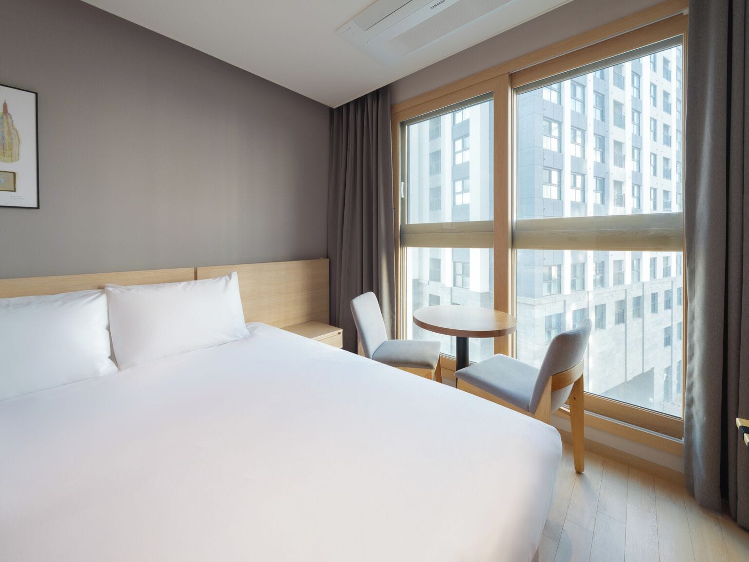 Quarto de hotel com uma cama grande, duas cadeiras, uma mesa pequena e vista para um edifício através de amplas janelas.