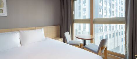 Quarto de hotel com uma cama grande, duas cadeiras, uma mesa pequena e vista para um edifício através de amplas janelas.