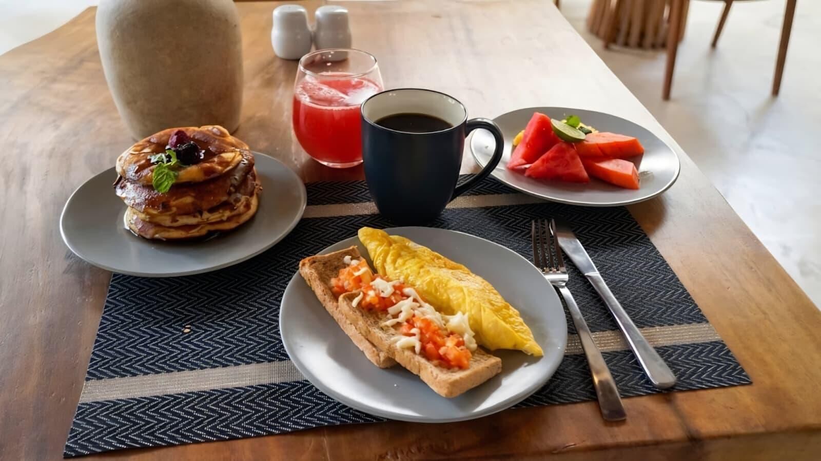 Disposição de café da manhã com panquecas, uma omelete e melancia em pratos, uma xícara de café e um copo de bebida vermelha.
