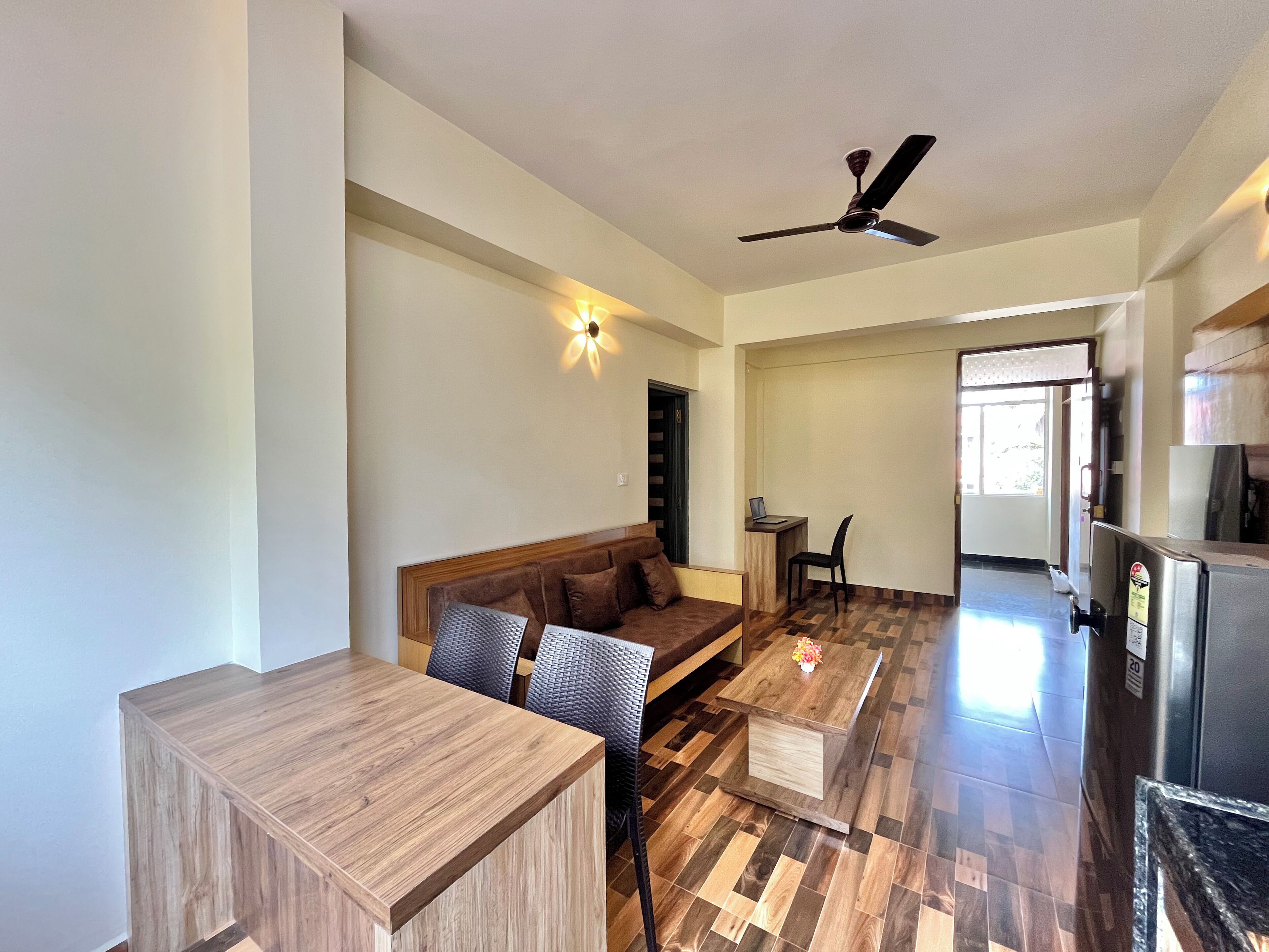 Uma sala moderna com uma mesa de centro de madeira, um sofá com almofadas, uma mesa de jantar e uma área de cozinha com uma geladeira.