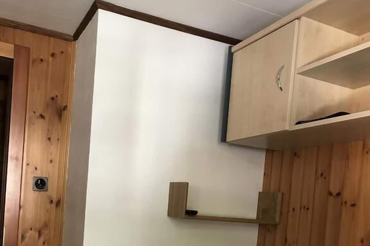 Uma cama de solteiro com roupa de cama branca e almofada preta, um guarda-roupa de madeira e uma pequena estante encostada a uma parede com painéis de madeira.