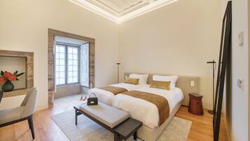 Quarto com cama grande, um banquinho, uma cadeira, mesa de cabeceira, abajur e janela.