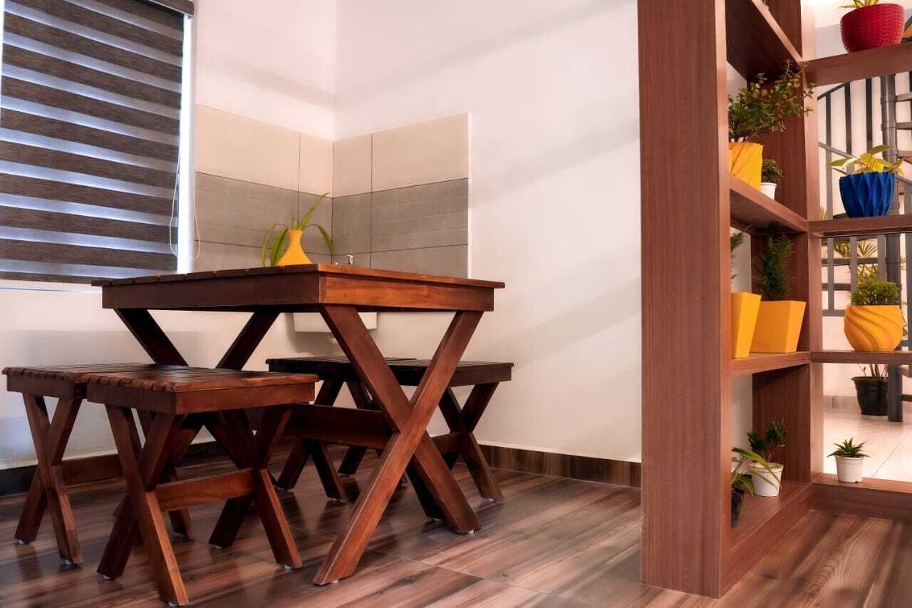 Uma área de refeições com mesa e cadeiras de madeira, uma estante com plantas em vasos e uma janela com persianas.